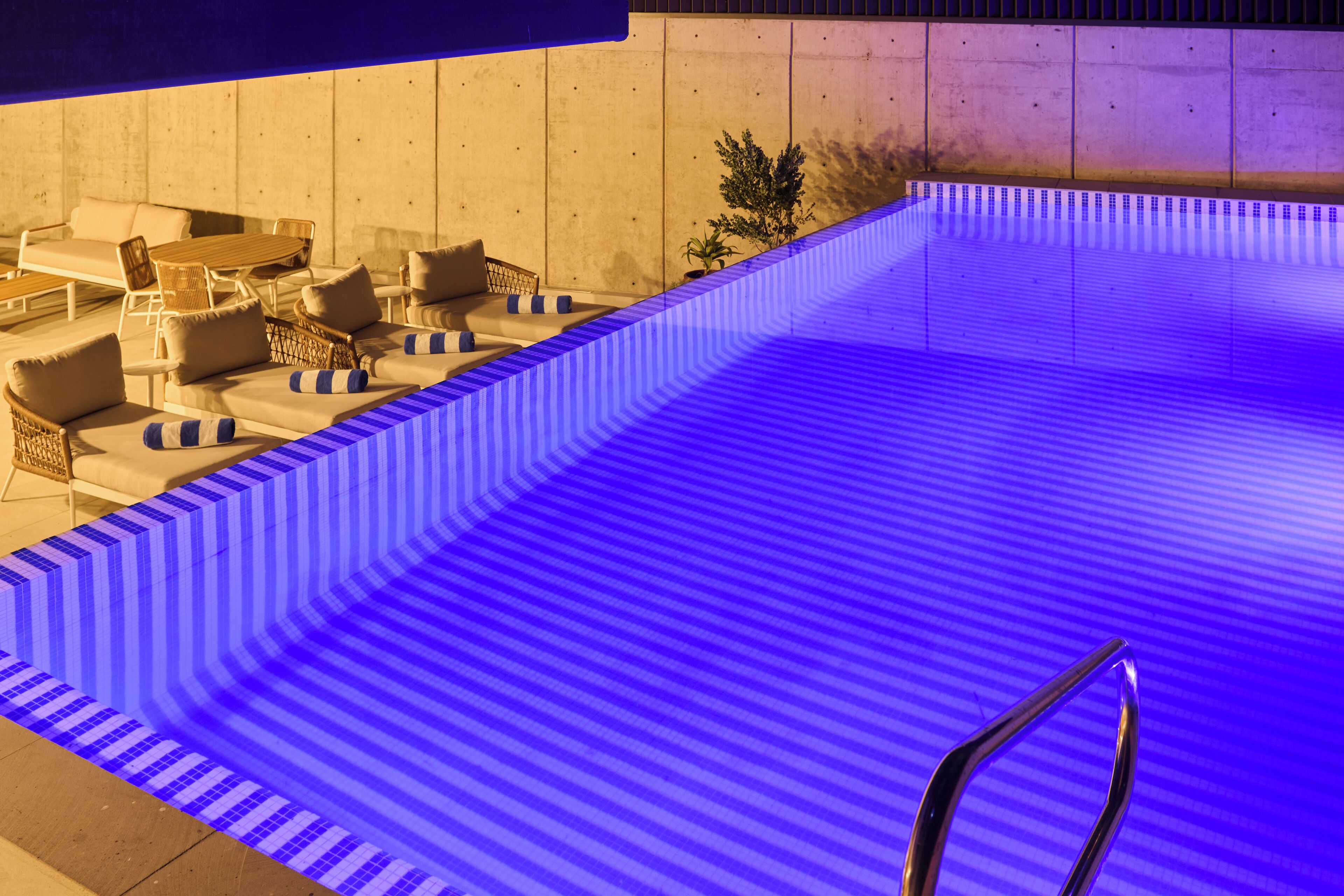 Piscina com iluminação azul, rodeada por mobiliário de vime e uma parede de concreto.