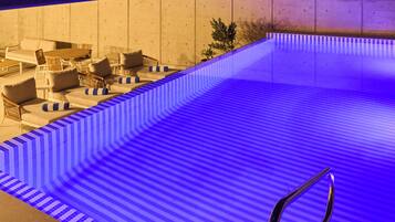 Piscina com iluminação azul, rodeada por mobiliário de vime e uma parede de concreto.