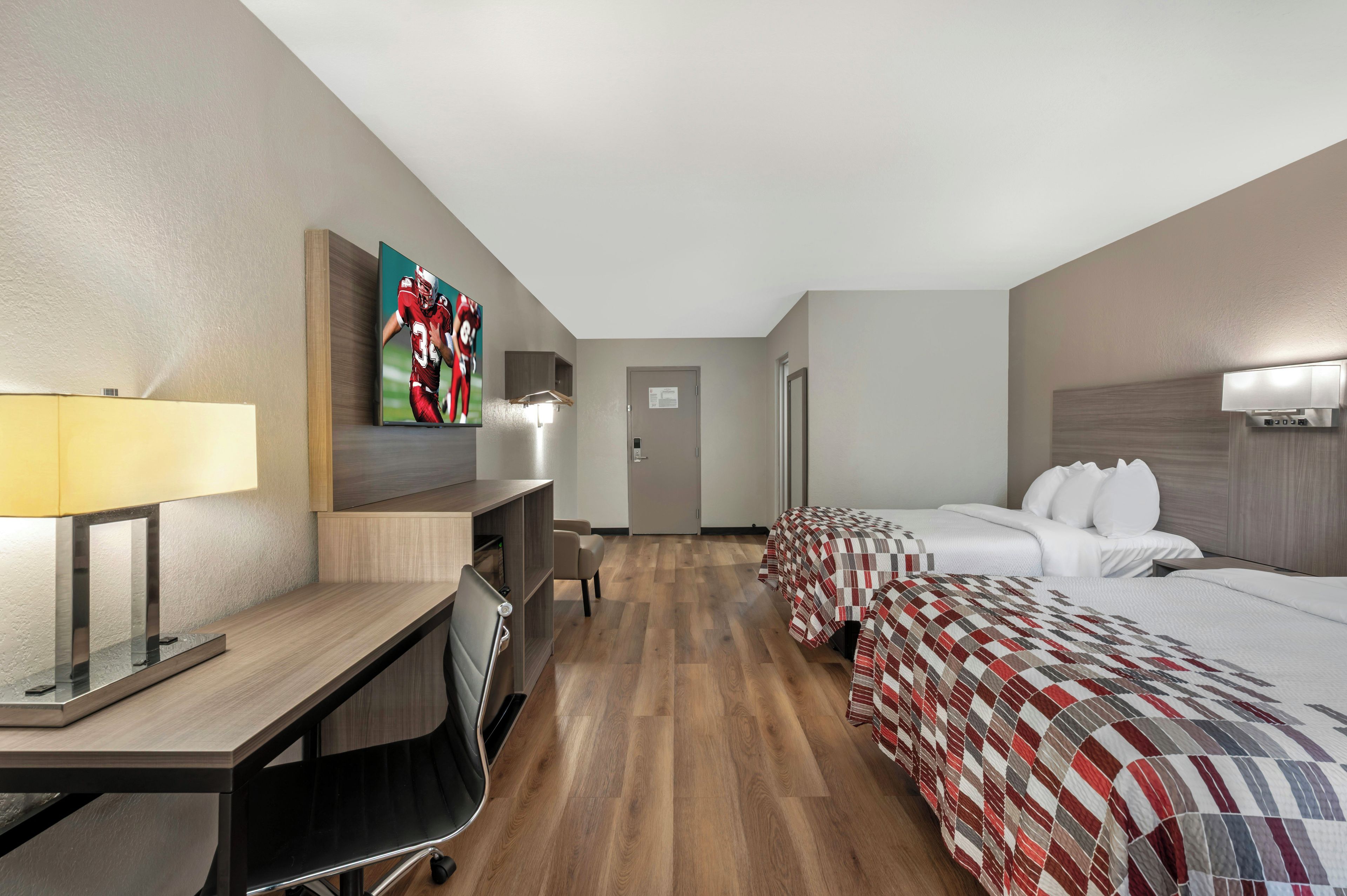 Quarto de hotel com uma escrivaninha, cama com colcha estampada, televisão presa na parede e porta para o corredor.