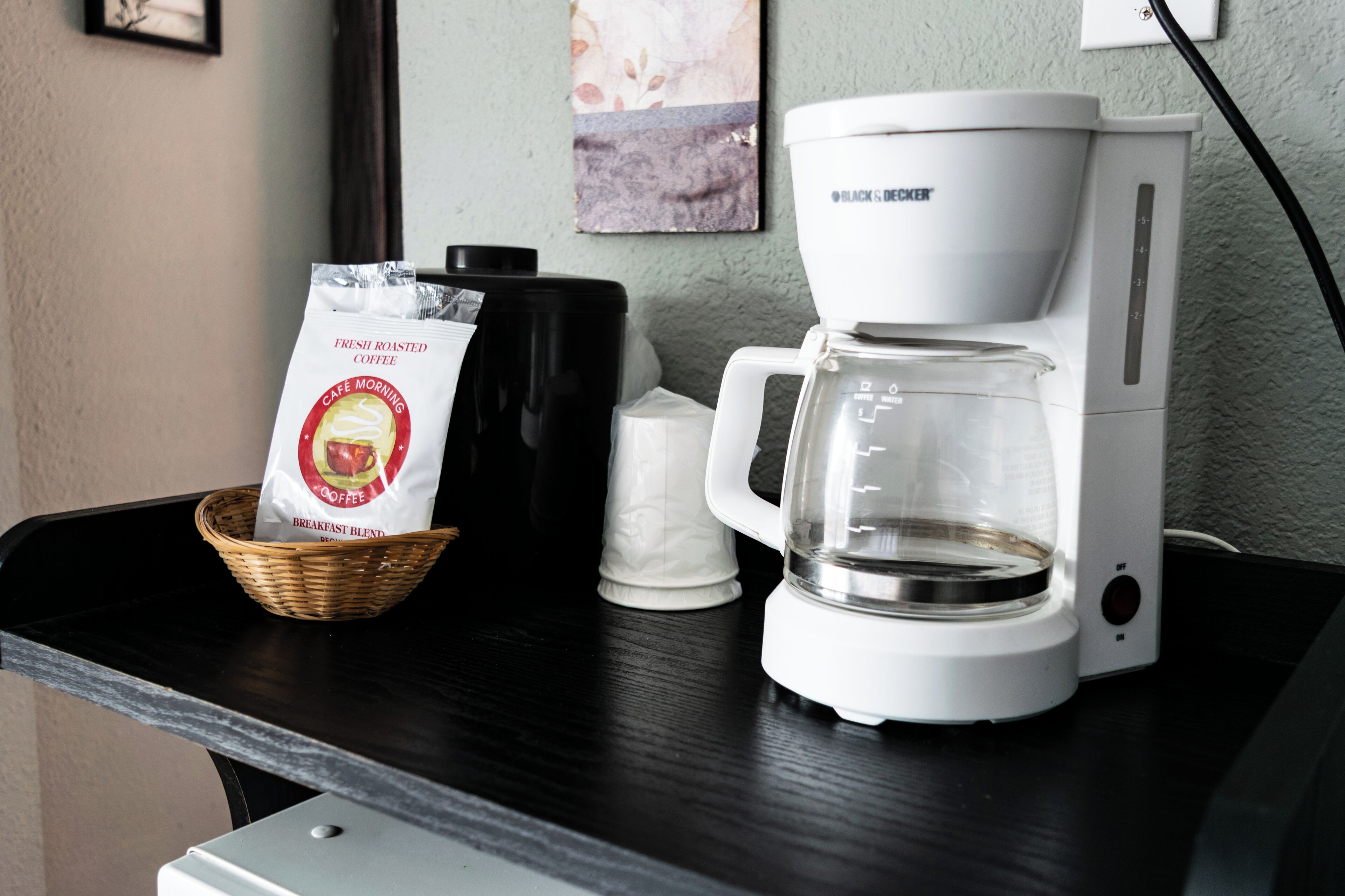 Uma cafeteira, uma cesta com um saco de café e uma jarra de vidro em uma bancada.