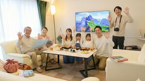 Um grupo de pessoas sentadas à volta de uma mesa, com comida e bebidas, num espaço com uma televisão grande exibindo uma paisagem montanhosa.