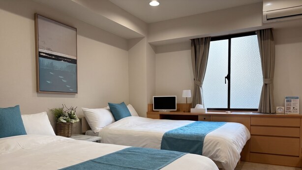 Quarto de hotel com duas camas, televisão, escrivaninha, janela com cortinas e ar-condicionado fixado na parede.