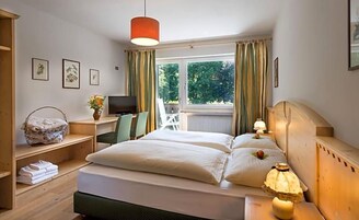 Quarto de hotel com cama, luminárias de cabeceira, escrivaninha com computador, cadeira, janela com cortinas e vista para o verde lá fora.