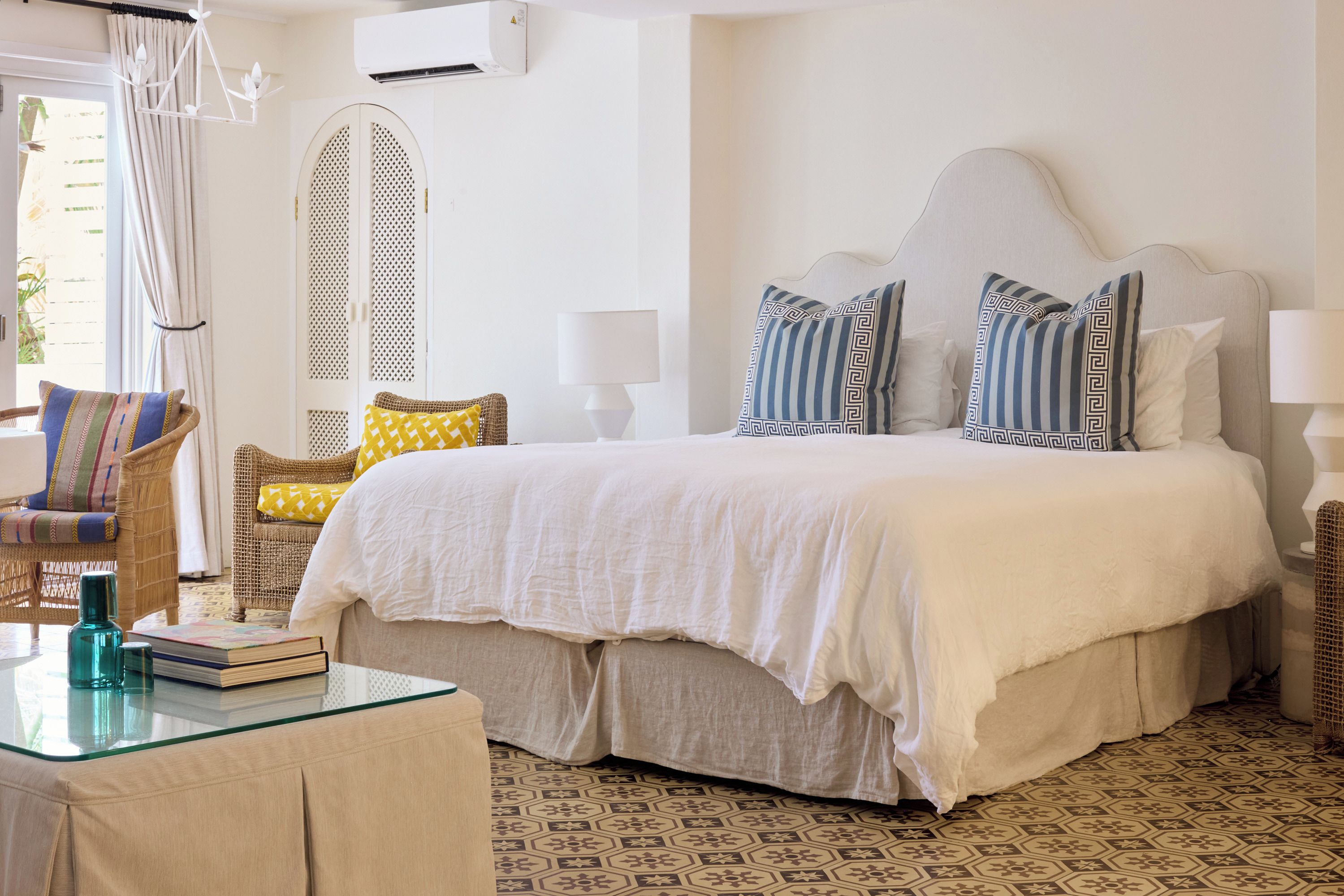 Quarto de hotel com uma cama grande, duas luminárias de cabeceira, uma cadeira de vime, uma mesa de centro de vidro e piso estampado.