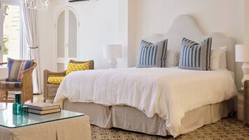 Quarto de hotel com uma cama grande, duas luminárias de cabeceira, uma cadeira de vime, uma mesa de centro de vidro e piso estampado.