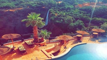 Uma área à beira da piscina com espreguiçadeiras, guarda-sóis e uma cascata ao fundo.