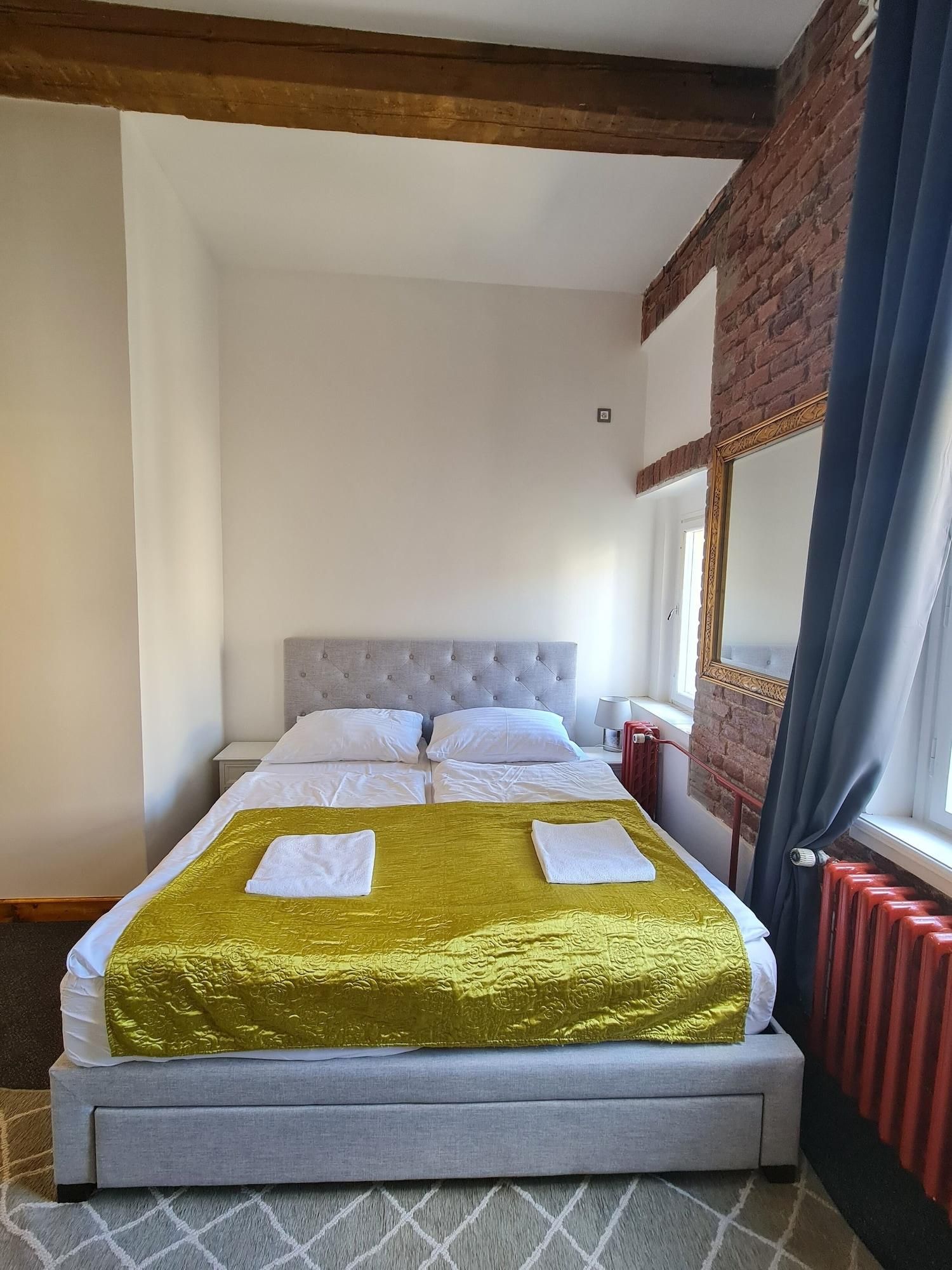 Quarto de hotel com cama, janela com cortinas, radiador e parede de tijolos.