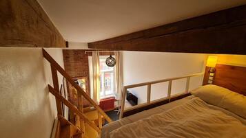 Quarto com escada de madeira, cama, mesa de cabeceira com abajur e vista para o exterior pela janela.
