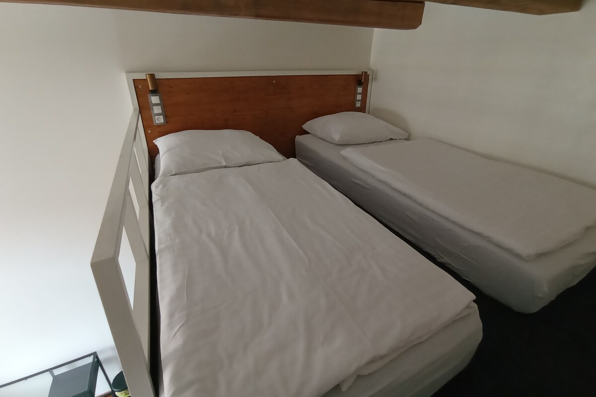 Quarto com duas camas de solteiro, cabeceira de madeira, roupa de cama branca e uma viga de madeira no teto.