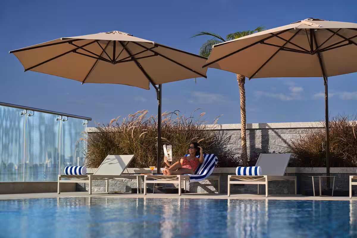Área à beira da piscina com espreguiçadeiras e guarda-sóis, uma pessoa lendo revista, e vista para o horizonte da cidade.