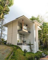 Uma casa moderna de dois andares com uma varanda no andar superior, rodeada por vegetação.