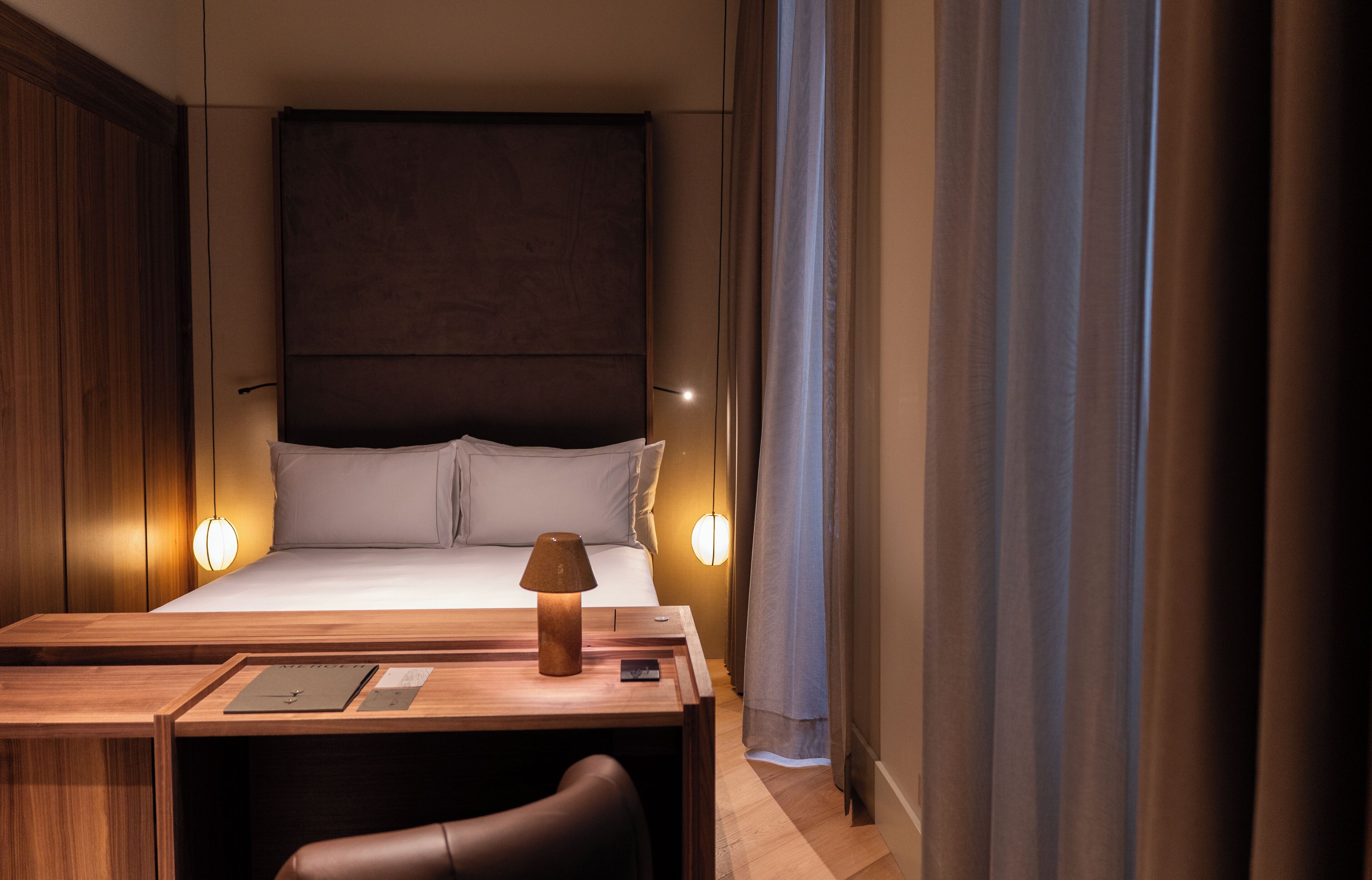 Quarto de hotel moderno com uma cama grande, abajures, uma escrivaninha de madeira e uma poltrona de couro.