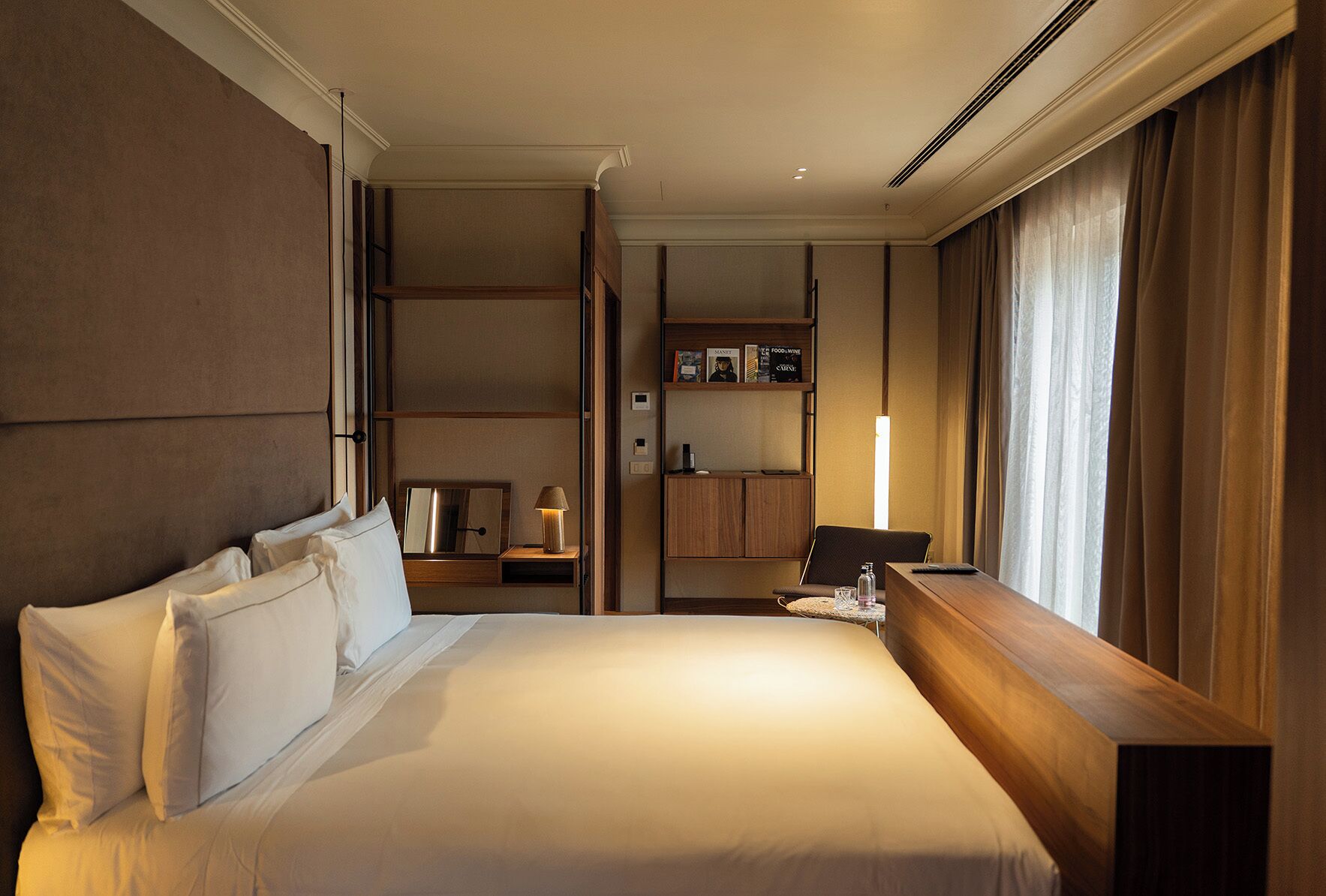 Quarto de hotel com uma cama grande, mesa de cabeceira com abajur, pequena mesa com garrafa, uma cadeira e um guarda-roupa.