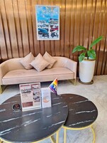 Uma área de recepção com um sofá, um vaso de planta, uma mesa com folhetos e um cartaz divulgando serviço de carro particular.