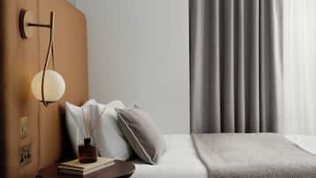 Uma cama bem arrumada com um cobertor cinzento, uma mesa de cabeceira com um livro e uma vela, e um abajur fixado na parede.