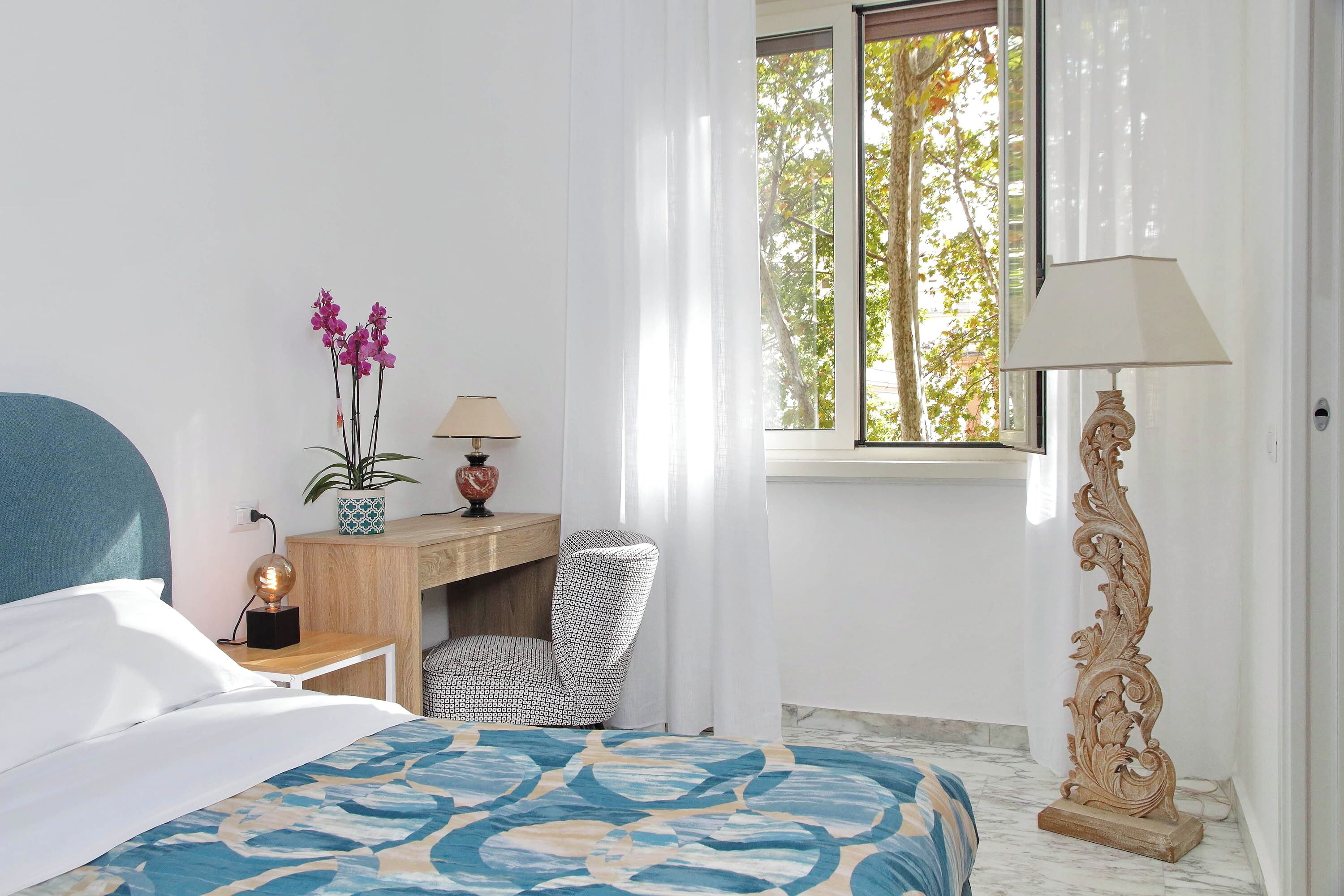 Quarto com cama, escrivaninha, cadeira, abajur e janela com vista para árvores.