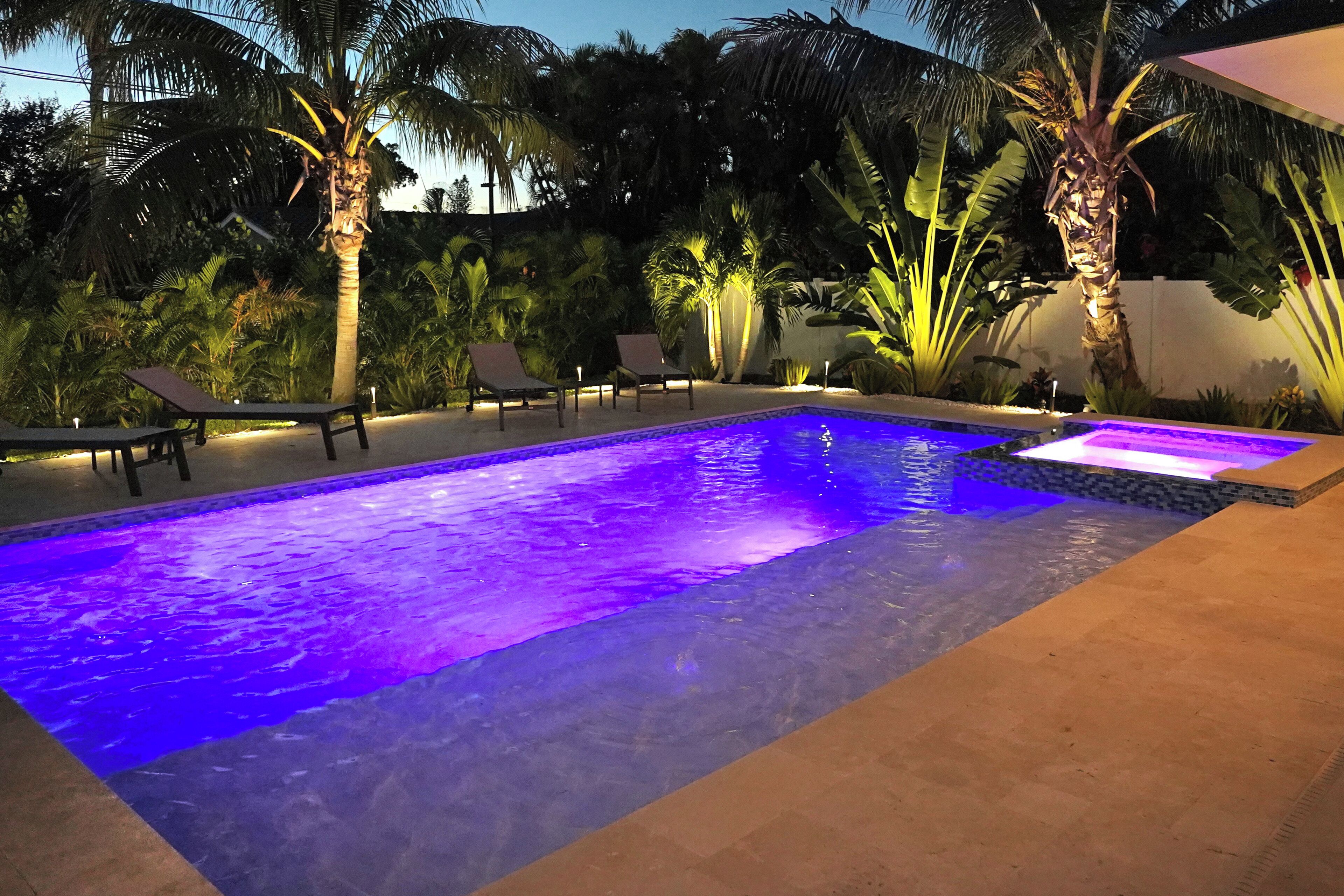 Piscina iluminada em azul, rodeada por palmeiras e espreguiçadeiras.
