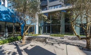 Um edifício moderno com grandes janelas de vidro, fachada azul e área ajardinada com árvores e assentos.