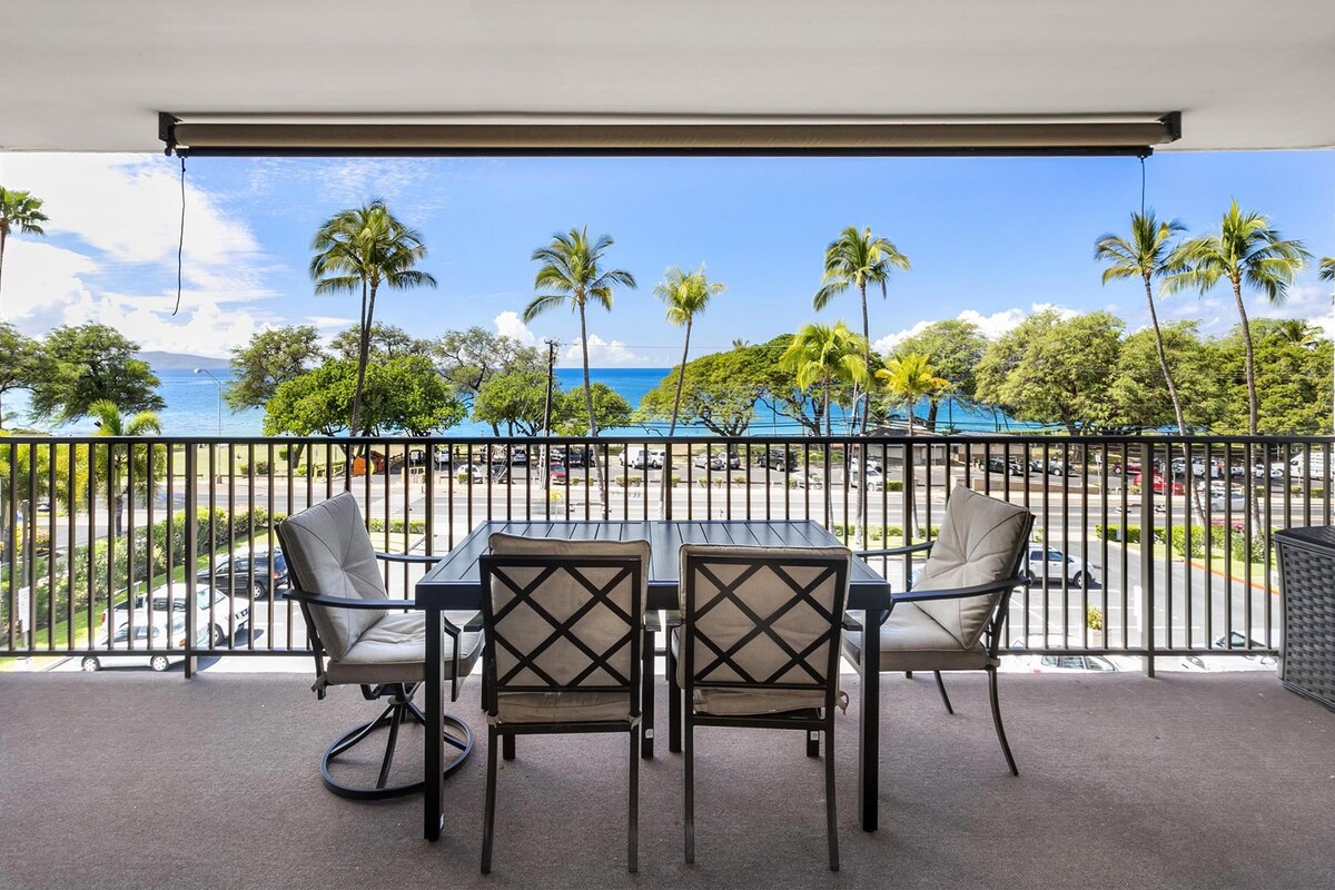 Varanda com mesa e cadeiras, vista para uma rua com palmeiras e o oceano.