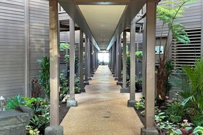 Uma galeria coberta com caminho de cascalho, ladeada por vegetação e colunas, que conduz a um edifício com placa indicando um hotel.