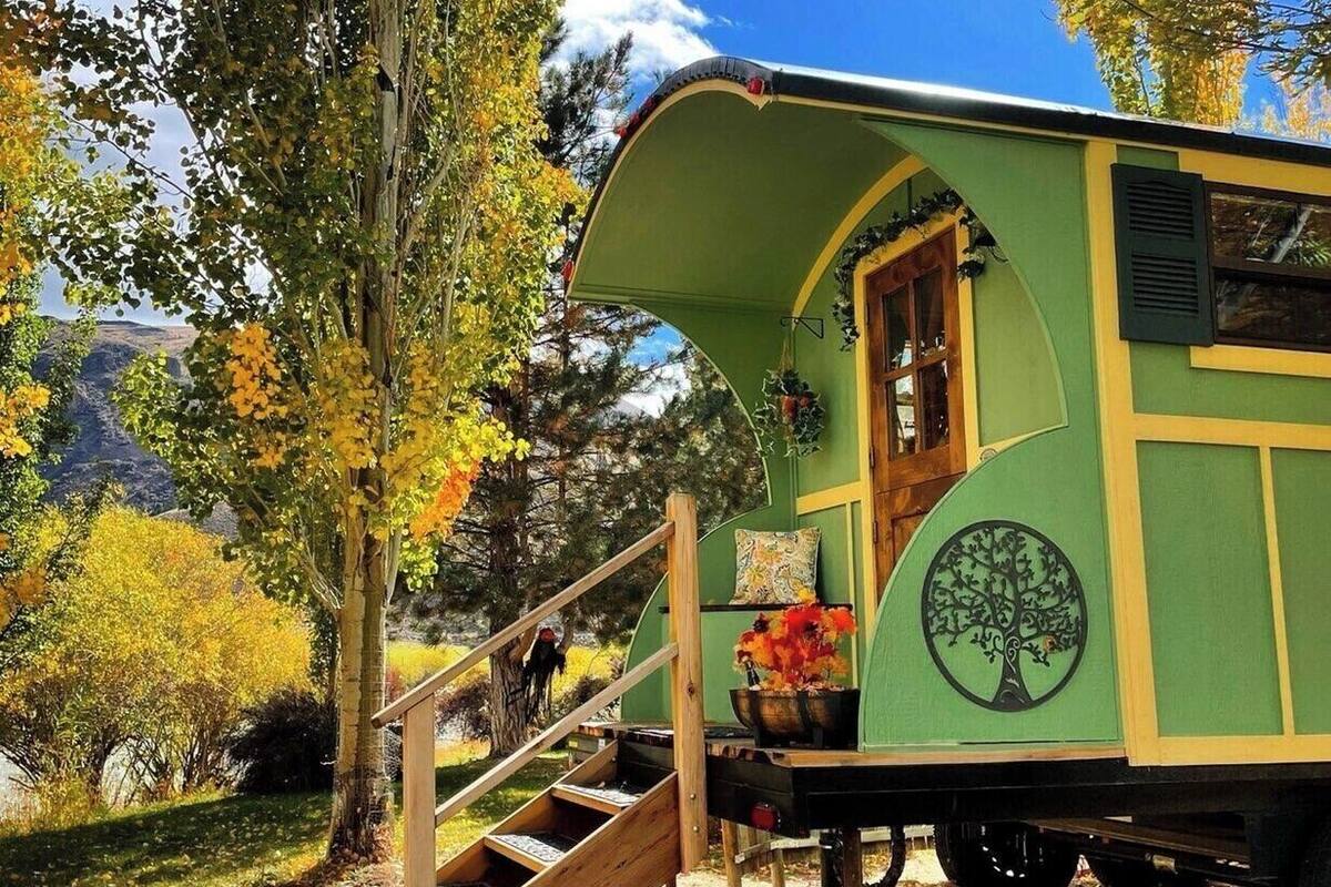 Um antigo vagão de trem verde com uma escadaria de madeira que leva à entrada, cercado por folhagem outonal e um cenário de montanhas.