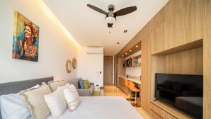 Quarto de hotel moderno com uma cama grande, ventilador de teto, TV de tela plana e área de cozinha ao fundo.