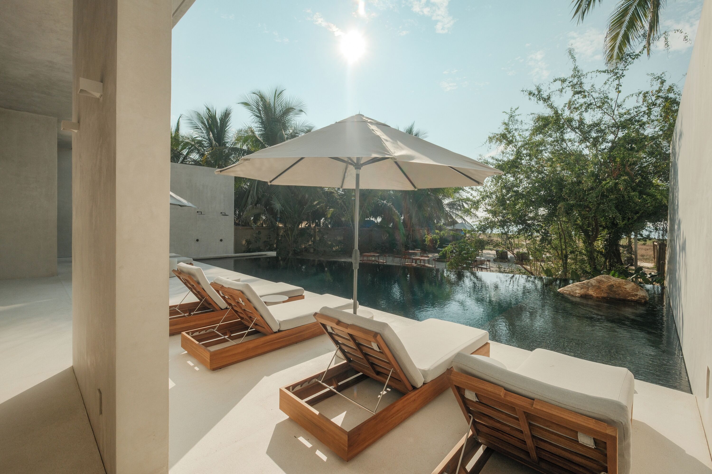 Área ao lado da piscina com espreguiçadeiras e um guarda-sol, cercada por palmeiras e céu claro.
