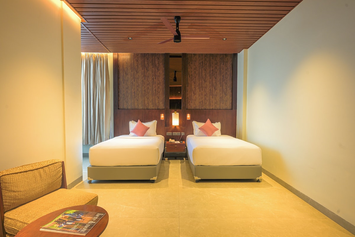 Quarto de hotel com duas camas, mesa de cabeceira, luminária, sofá e uma revista sobre a mesa.