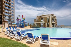 Área de piscina com espreguiçadeiras, parquinho infantil e um edifício com varandas.