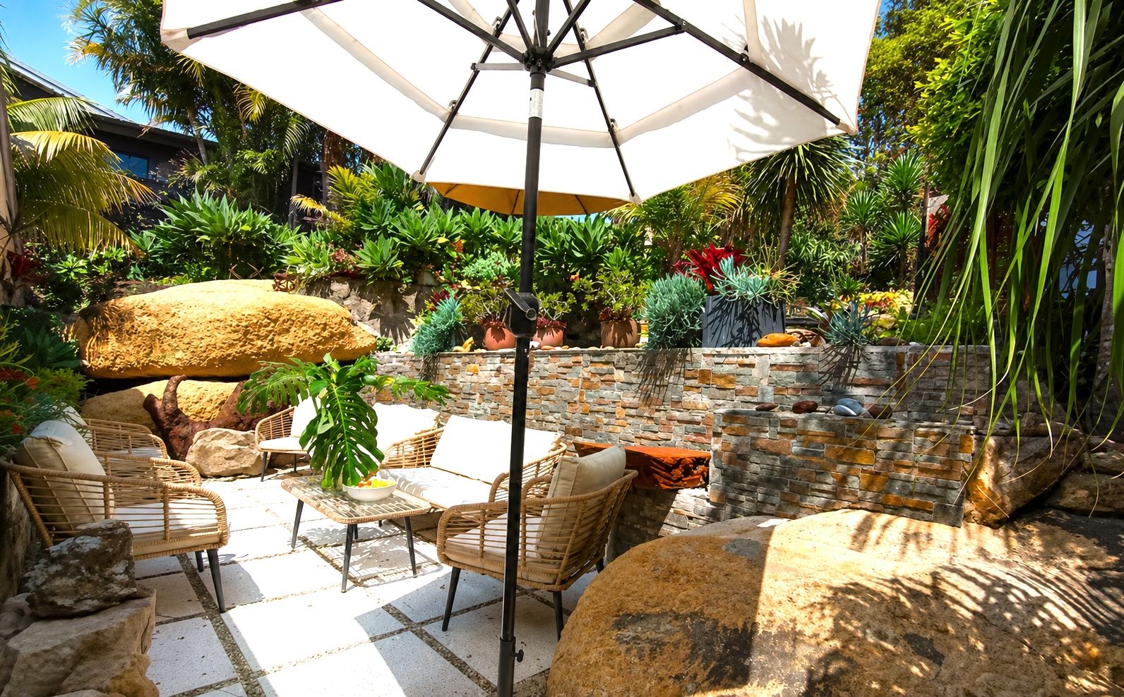 Área de terraço ao ar livre com móveis de vime, parede de pedra e diversos vasos de plantas.