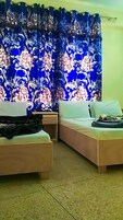 Quarto com duas camas de madeira, uma mesinha de cabeceira e cortina estampada com flores.