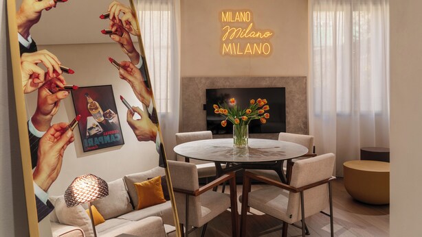 Uma sala de estar moderna com uma mesa de jantar redonda, sofás bege, uma TV e um letreiro neon com a inscrição "MILANO MILANO".
