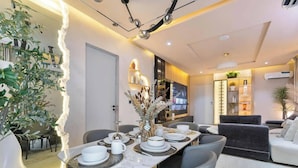 Uma sala de jantar moderna com uma mesa comprida posta para quatro pessoas, rodeada por cadeiras cinzentas. Há uma televisão montada na parede e uma estante com objetos decorativos.