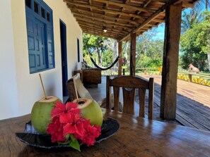 Uma mesa de madeira com dois cocos e flores vermelhas, uma rede e uma cadeira de madeira em área externa coberta, com vista para o verde.