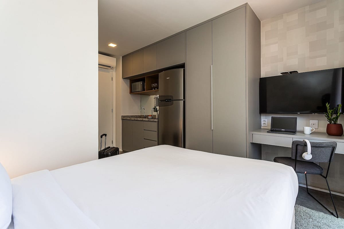 Quarto de hotel moderno com cama, cozinha compacta, televisão de tela plana, escrivaninha com laptop e uma cadeira.