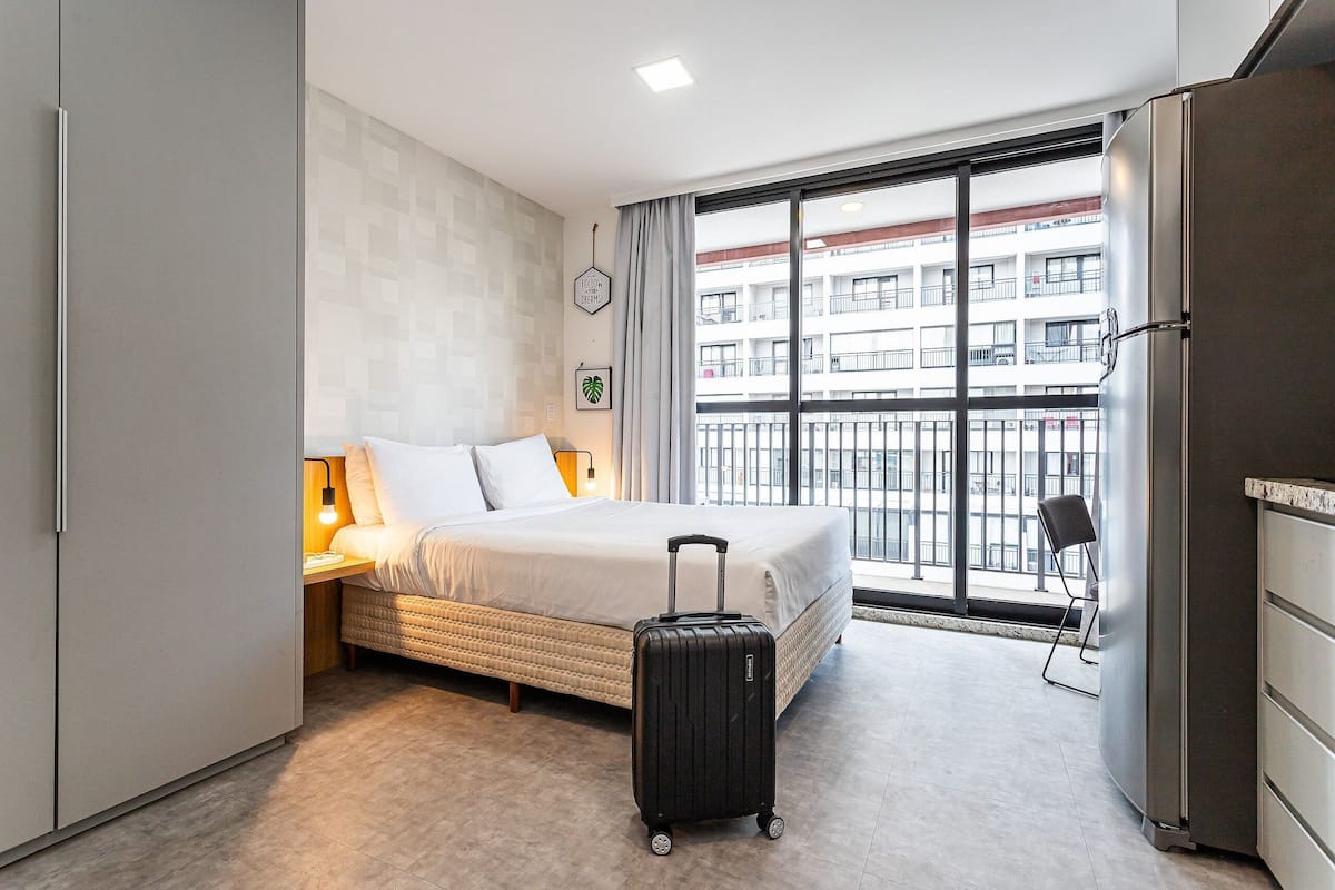 Quarto de hotel com cama, mala, escrivaninha com luminária, cadeira e janela grande com vista para um edifício.