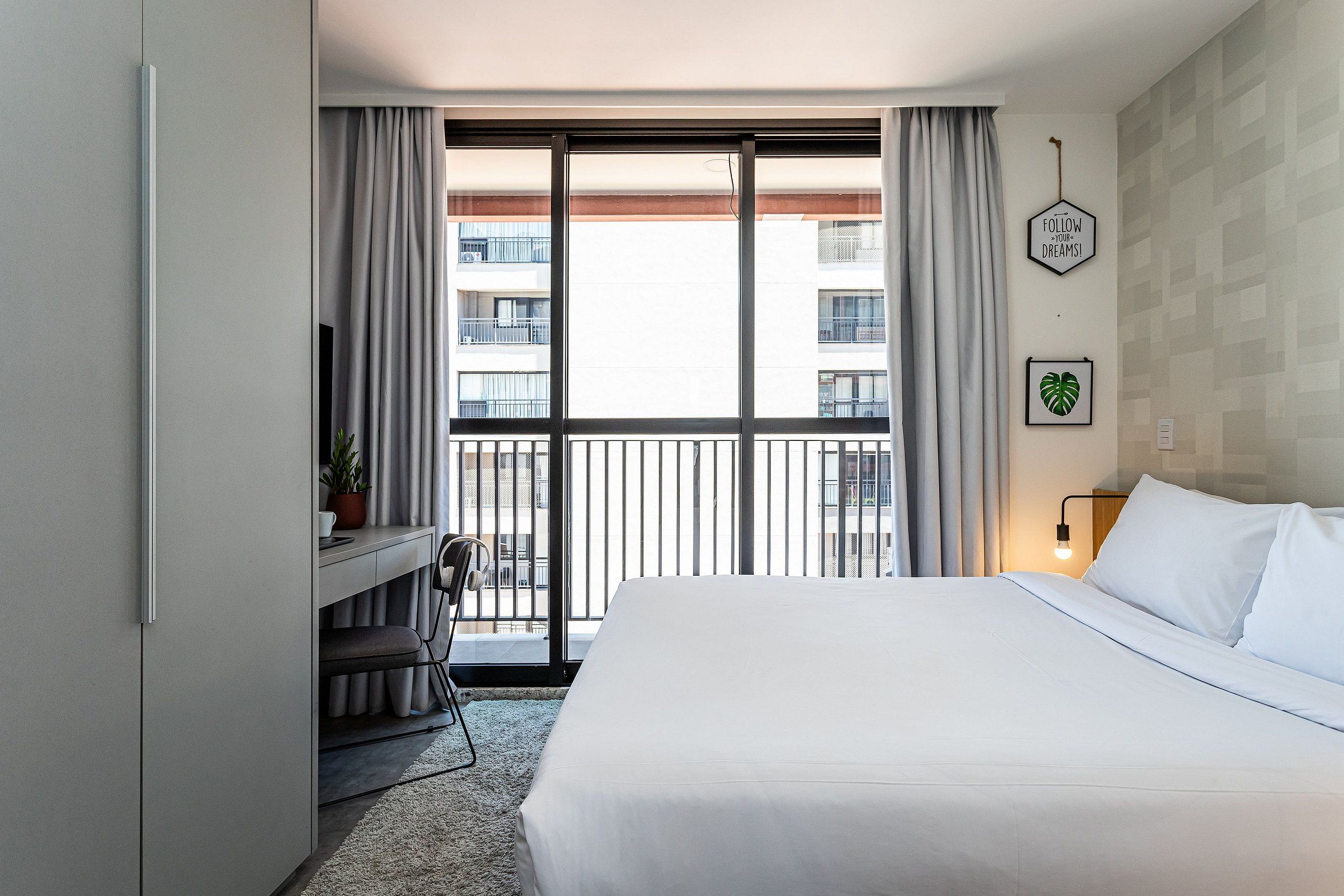 Quarto de hotel com uma cama, uma escrivaninha com cadeira, um vaso de planta, uma janela com cortinas, uma luminária na parede e um quadro emoldurado.