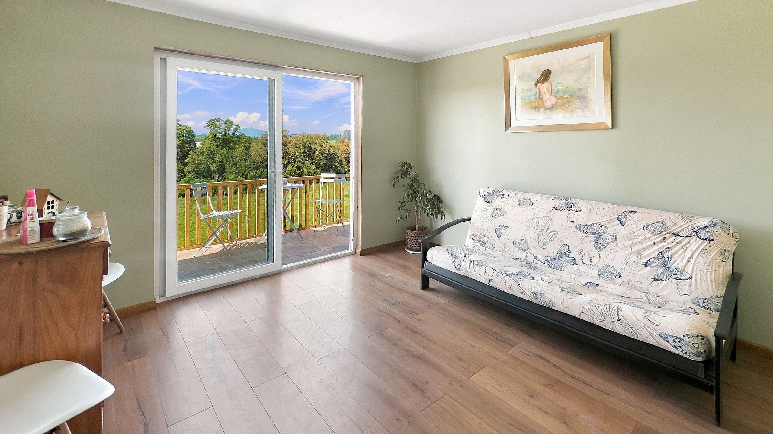 Quarto com piso de madeira, sofá estampado com motivos florais, mesa de jantar com cadeiras, pintura na parede e porta de vidro deslizante que dá para uma área externa.