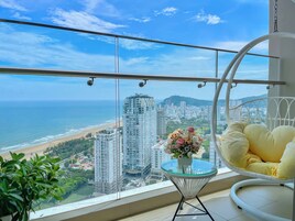 Uma varanda com vista desobstruída para o oceano e a paisagem urbana, uma cadeira de vime e uma pequena mesa com um vaso de flores.