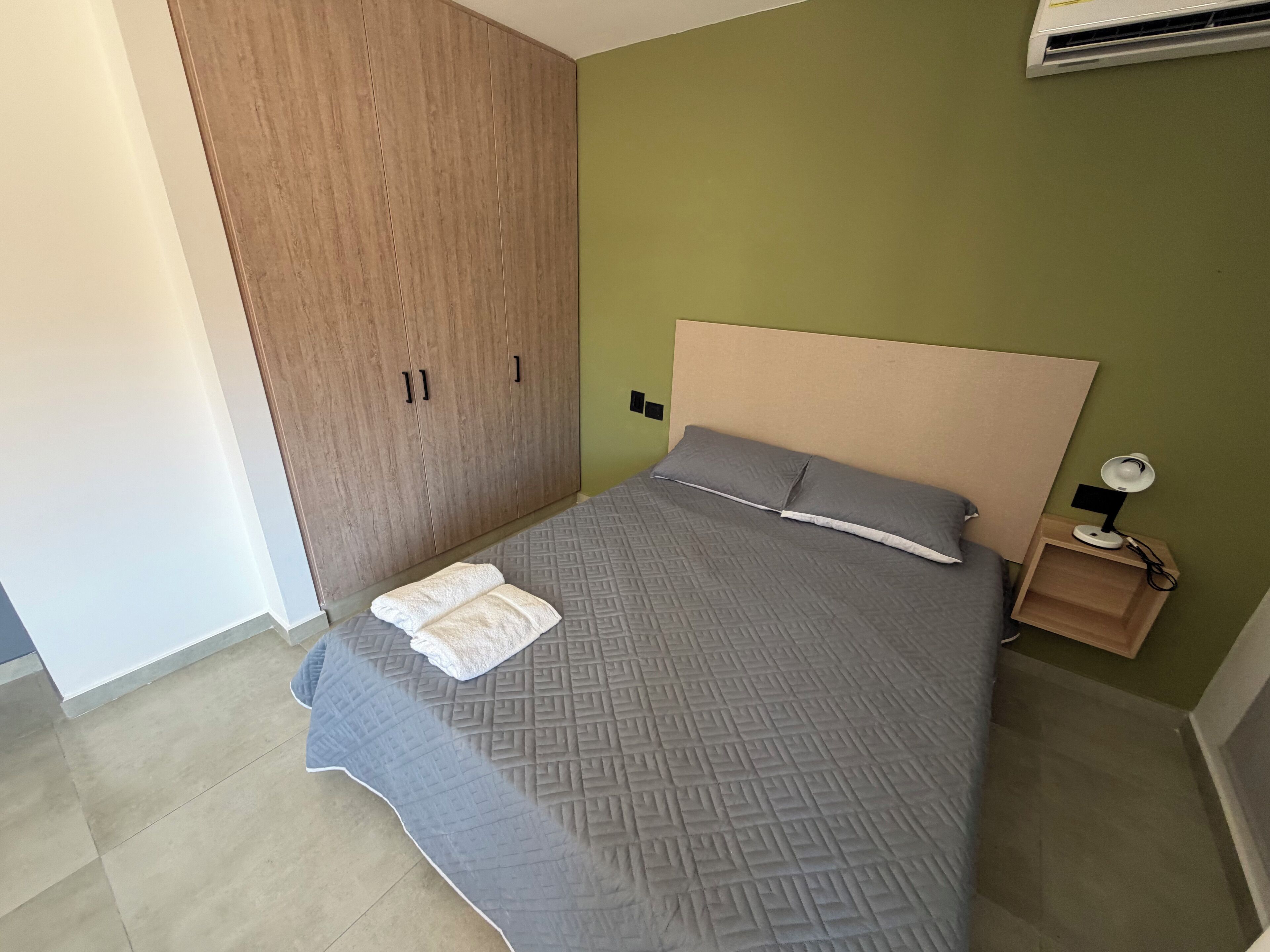Quarto com cama, mesa de cabeceira, guarda-roupa de madeira e ar condicionado.