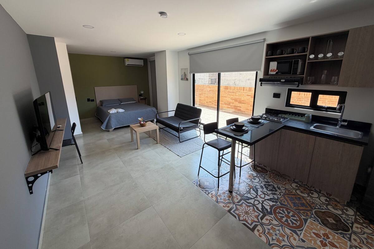 Quarto de hotel moderno com cama, escrivaninha, cadeira, televisão, espaço de cozinha e uma janela grande.