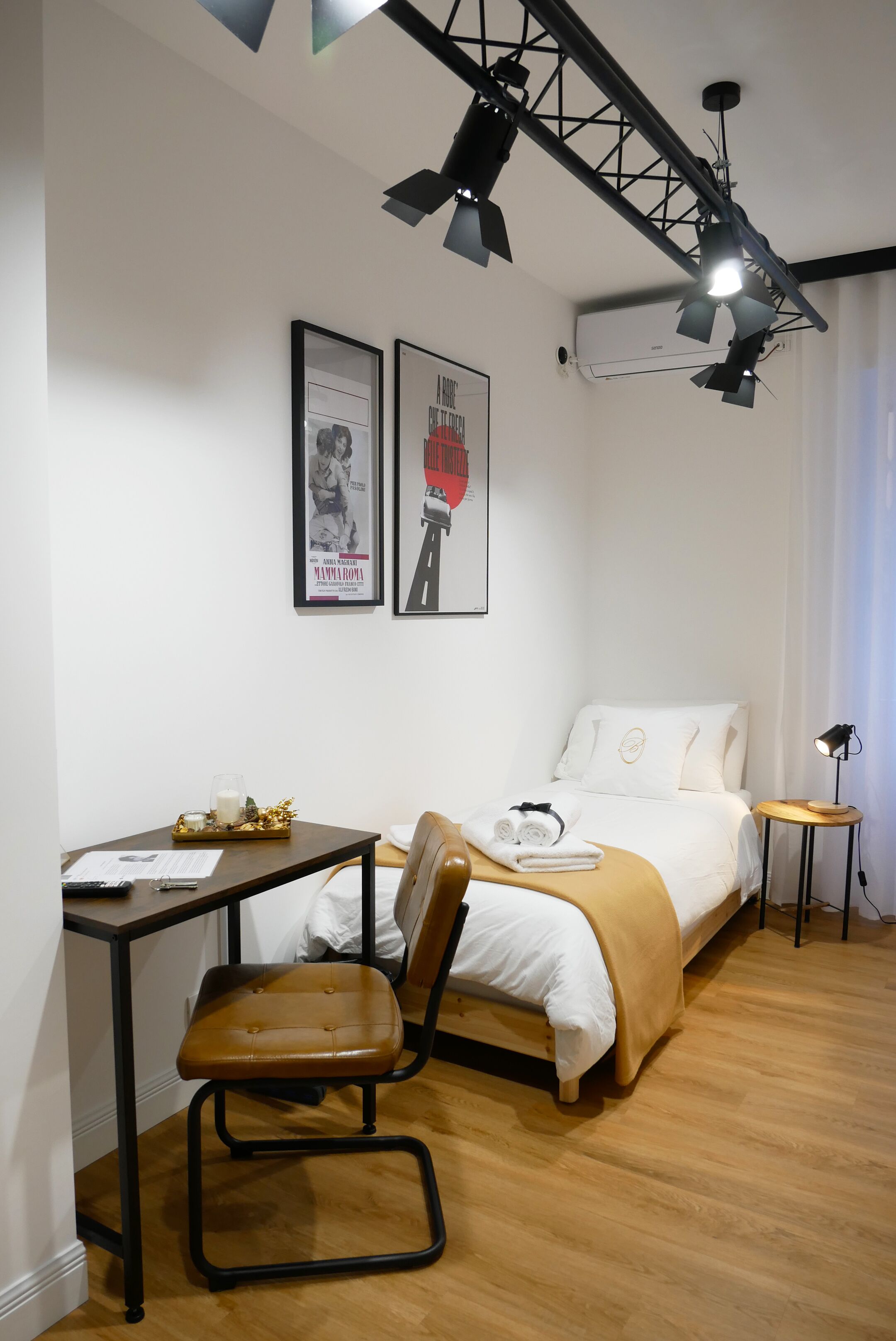 Quarto de hotel bem arrumado com uma cama, uma mesa de trabalho e uma cadeira. Há duas quadros na parede, uma mesinha com uma vela e um abajur em uma mesa lateral.