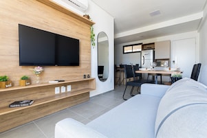 Uma sala de estar moderna com uma televisão de tela plana, estantes de madeira, um sofá e um espaço de jantar com cadeiras e uma mesa.
