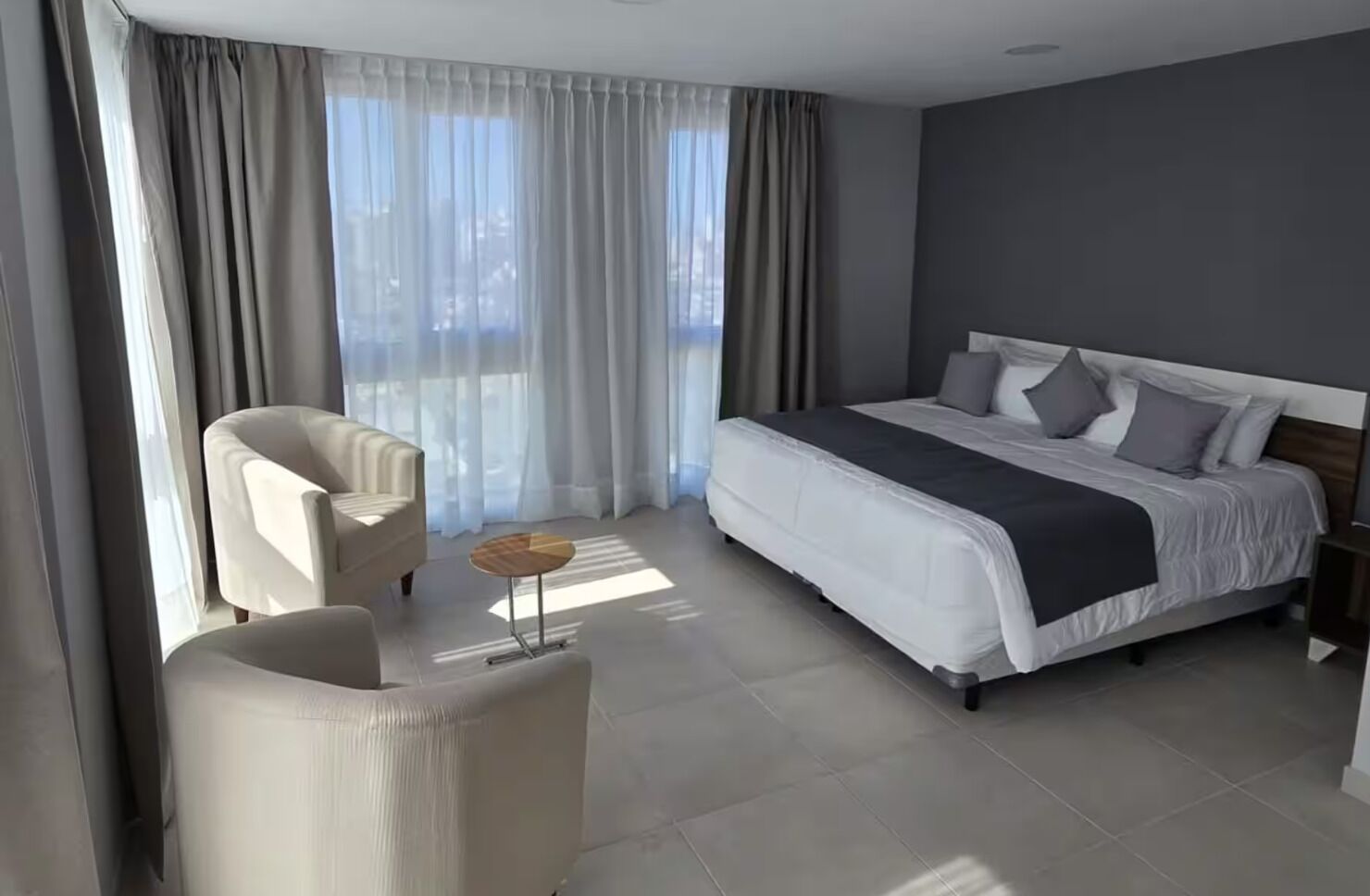 Quarto de hotel moderno com uma cama grande, uma cadeira, uma pequena mesa e vista através de cortinas leves.