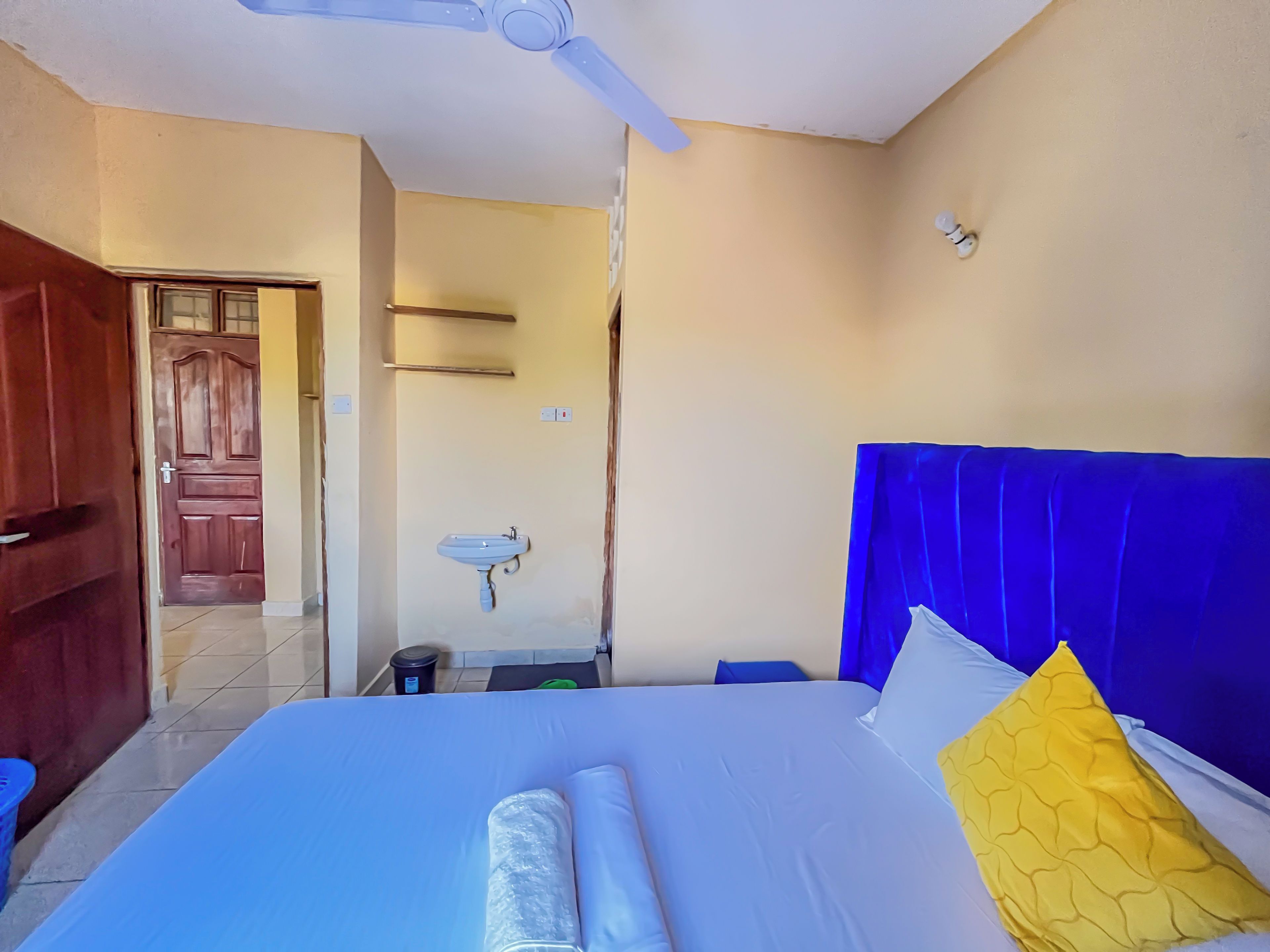 Quarto com cama, ventilador de teto, banheiro com pia e porta para outro cômodo.