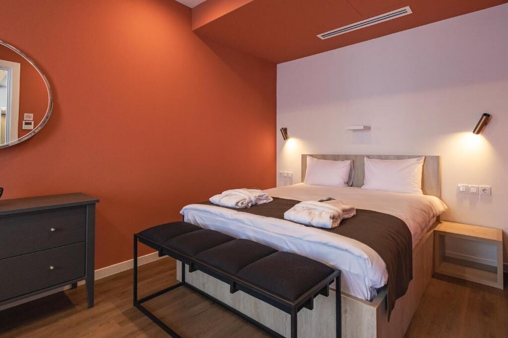 Quarto com cama, mesas de cabeceira, um banquinho, cômoda e espelho.
