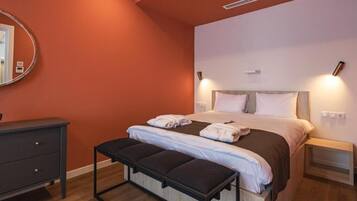 Quarto com cama, mesas de cabeceira, um banquinho, cômoda e espelho.