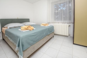 Cama bem arrumada com um cobertor azul e duas almofadas, janela com cortinas leves e um radiador na parede.
