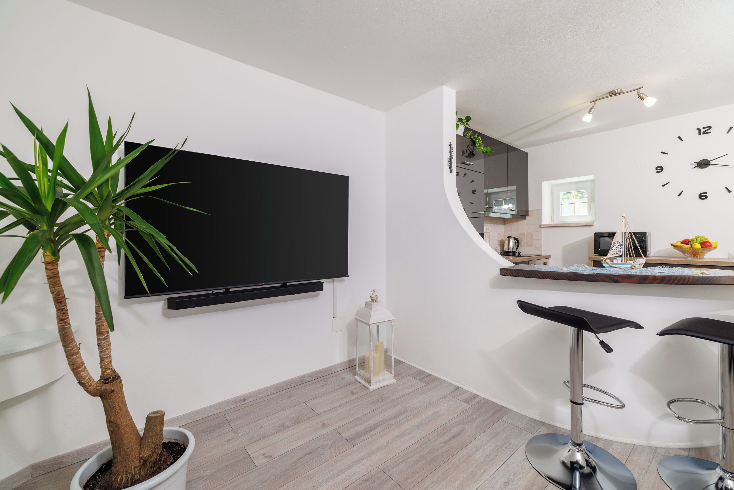 Uma sala de estar moderna com uma TV de tela plana, um vaso de planta, um lança branca e uma área de cozinha com um relógio e banquetas de bar.