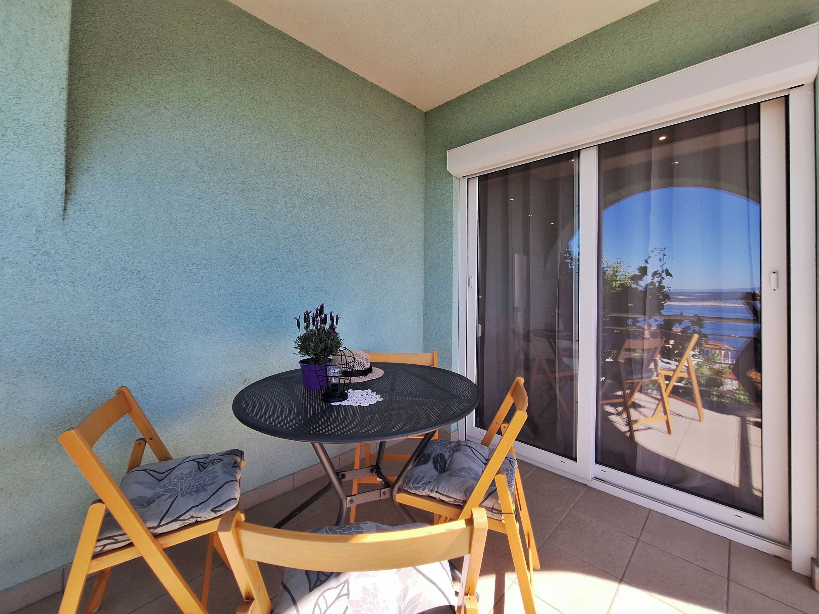 Uma varanda com mesa e cadeiras, um vaso de planta e vista para o mar.
